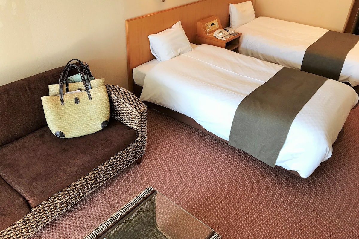 Quarto de hotel com duas camas, um sofá marrom e uma mesinha com telefone e luminária.
