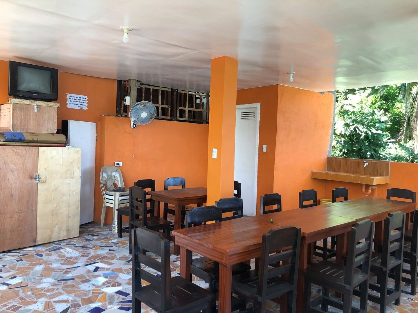 Quarto com paredes laranja, mesas e cadeiras de madeira, um ventilador de teto e uma televisão fixada na parede.