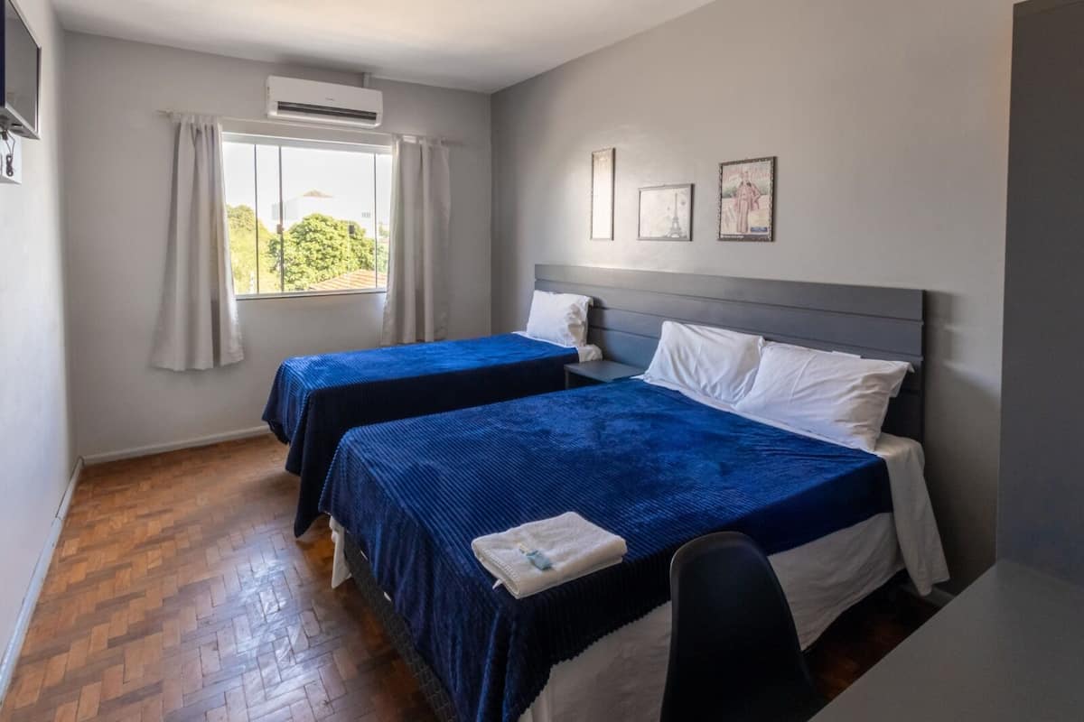 Quarto de hotel com duas camas, janela com cortinas, ar condicionado e uma pequena mesa com uma cadeira.