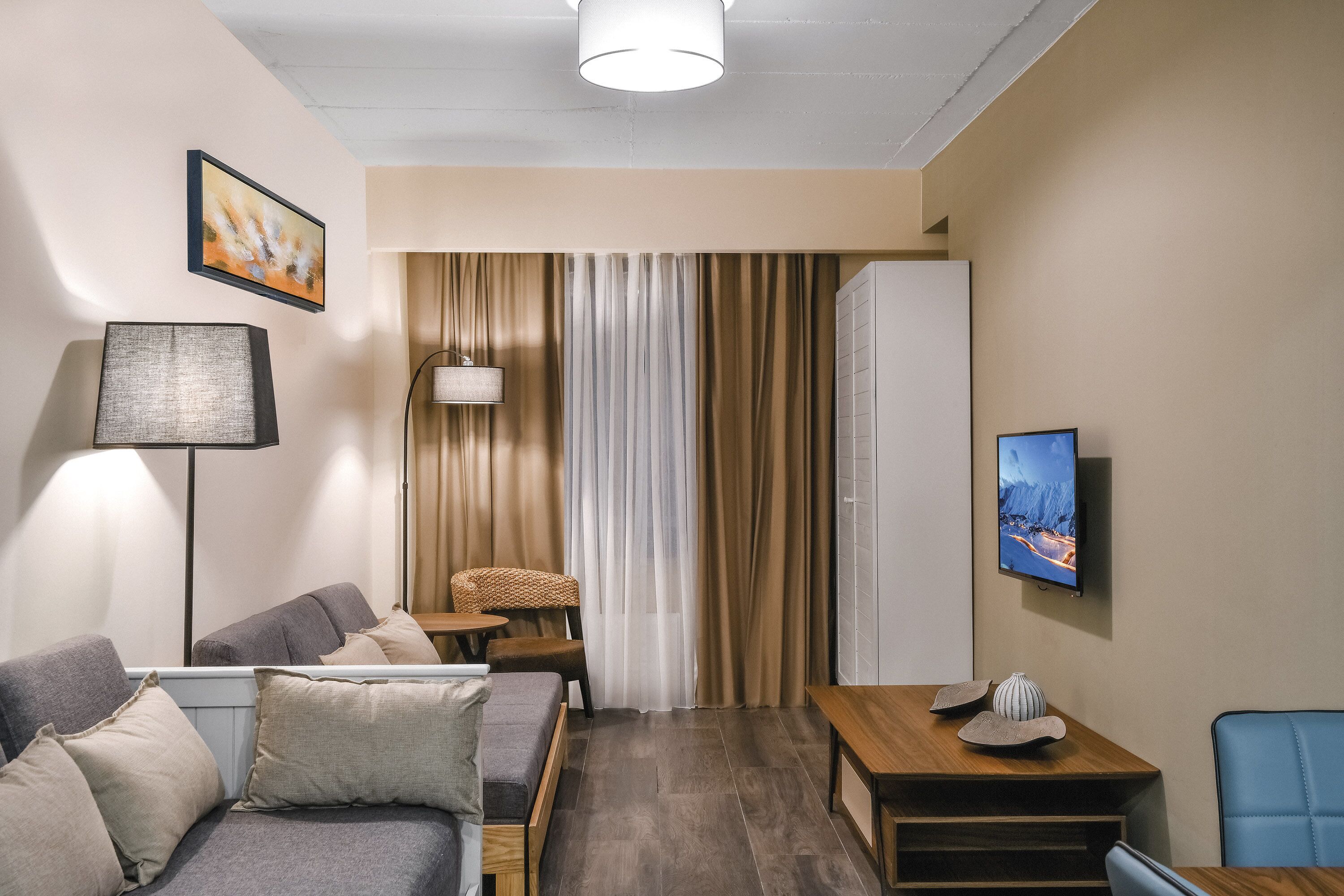 Um quarto moderno e bem iluminado, com um sofá, uma mesa pequena, uma cadeira, uma luminária e uma TV.