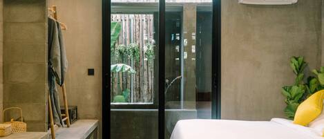 Quarto moderno com cama, um banquinho e vista para um jardim através de uma porta de vidro.