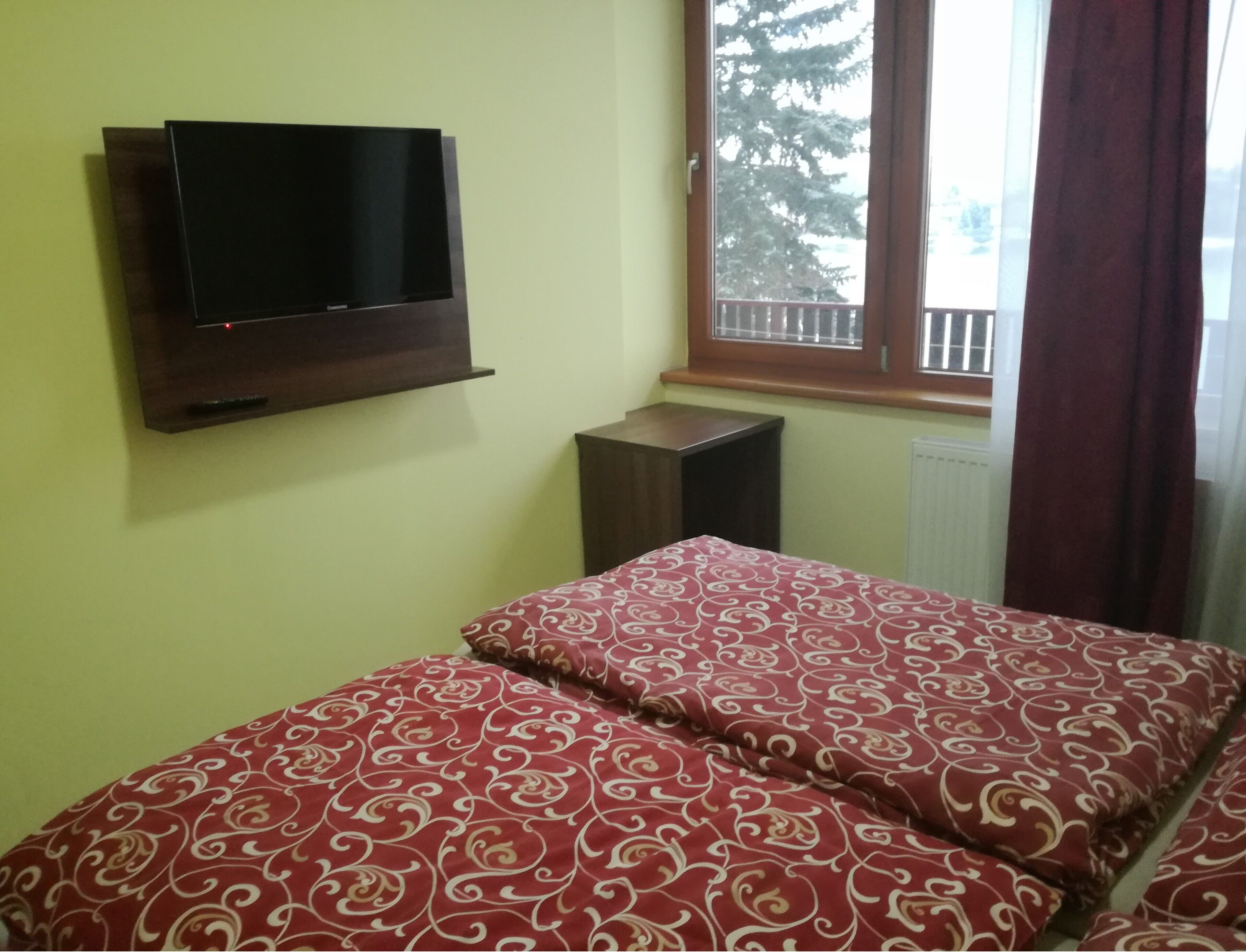 Quarto com cama, televisão na parede, mesinha e janela com vista para árvores.