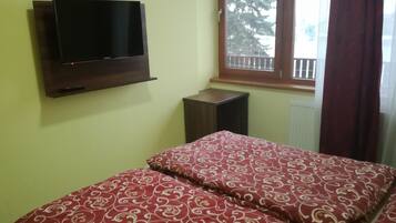 Quarto com cama, televisão na parede, mesinha e janela com vista para árvores.
