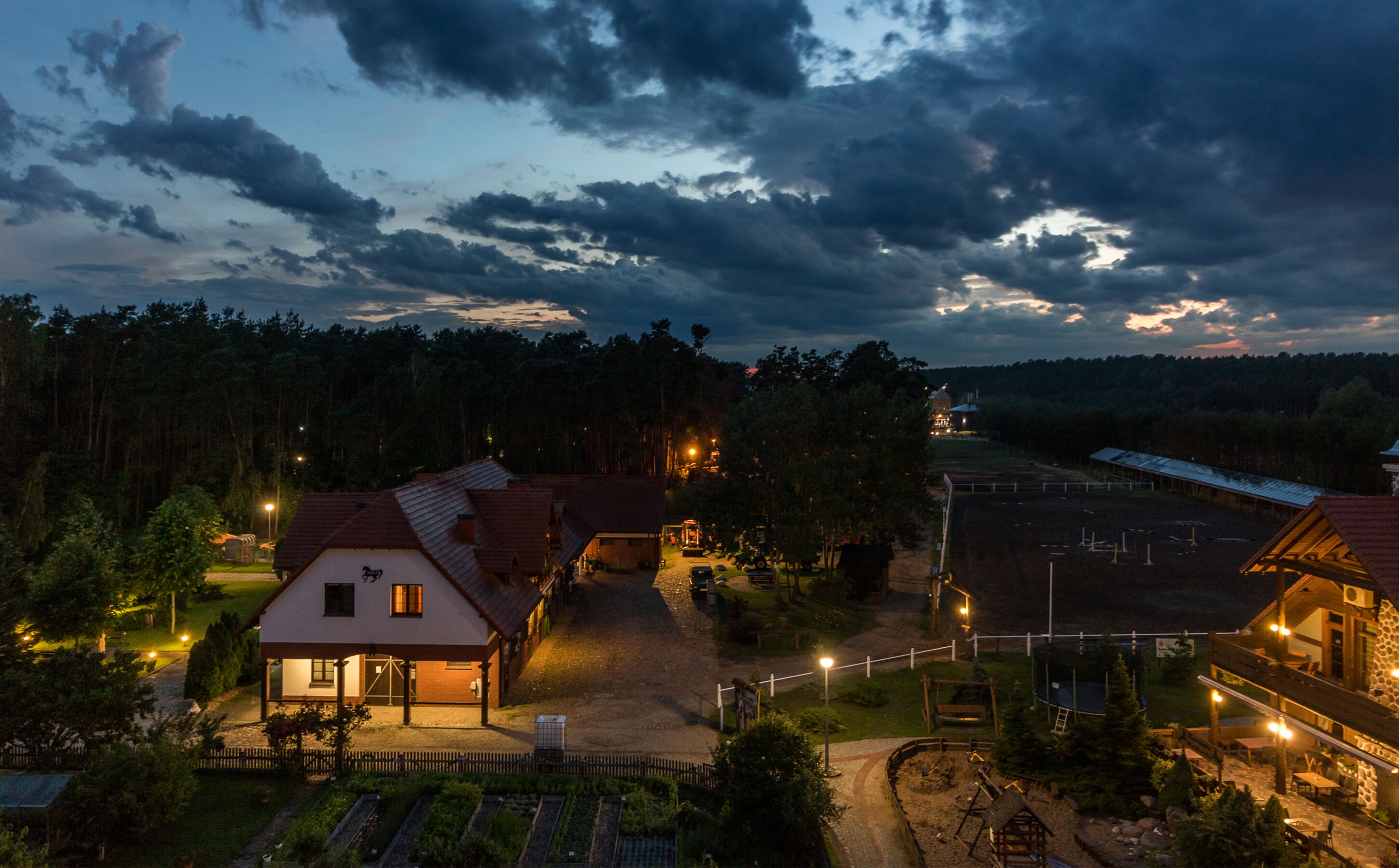 Cenário rural com um edifício principal, tendo um alpendre amplo e uma construção menor anexa. Vários edifícios menores e uma área cercada com um parquinho.