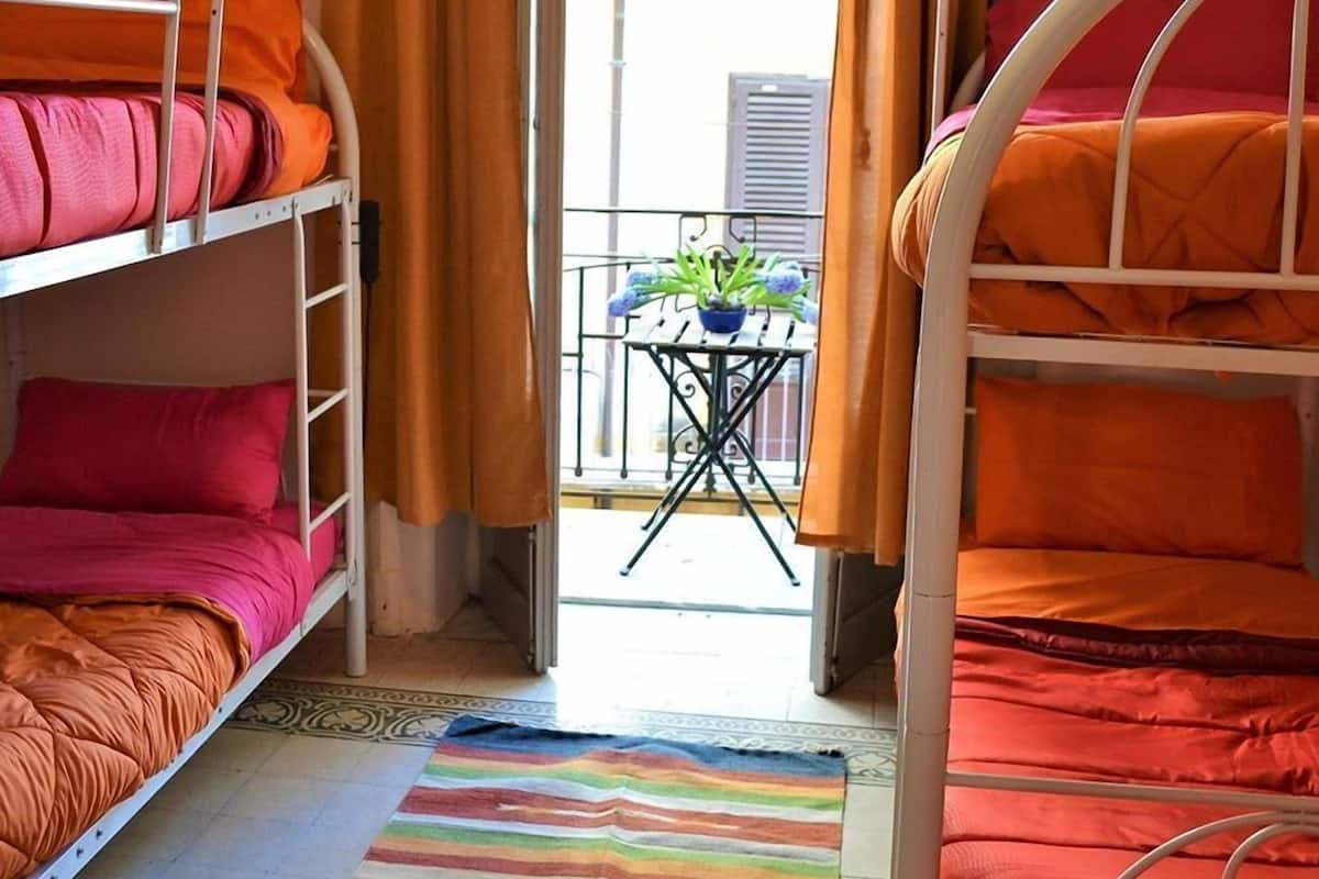Quarto tipo dormitório com beliches, uma pequena varanda com mesa e planta em vaso, e um tapete listrado e colorido.