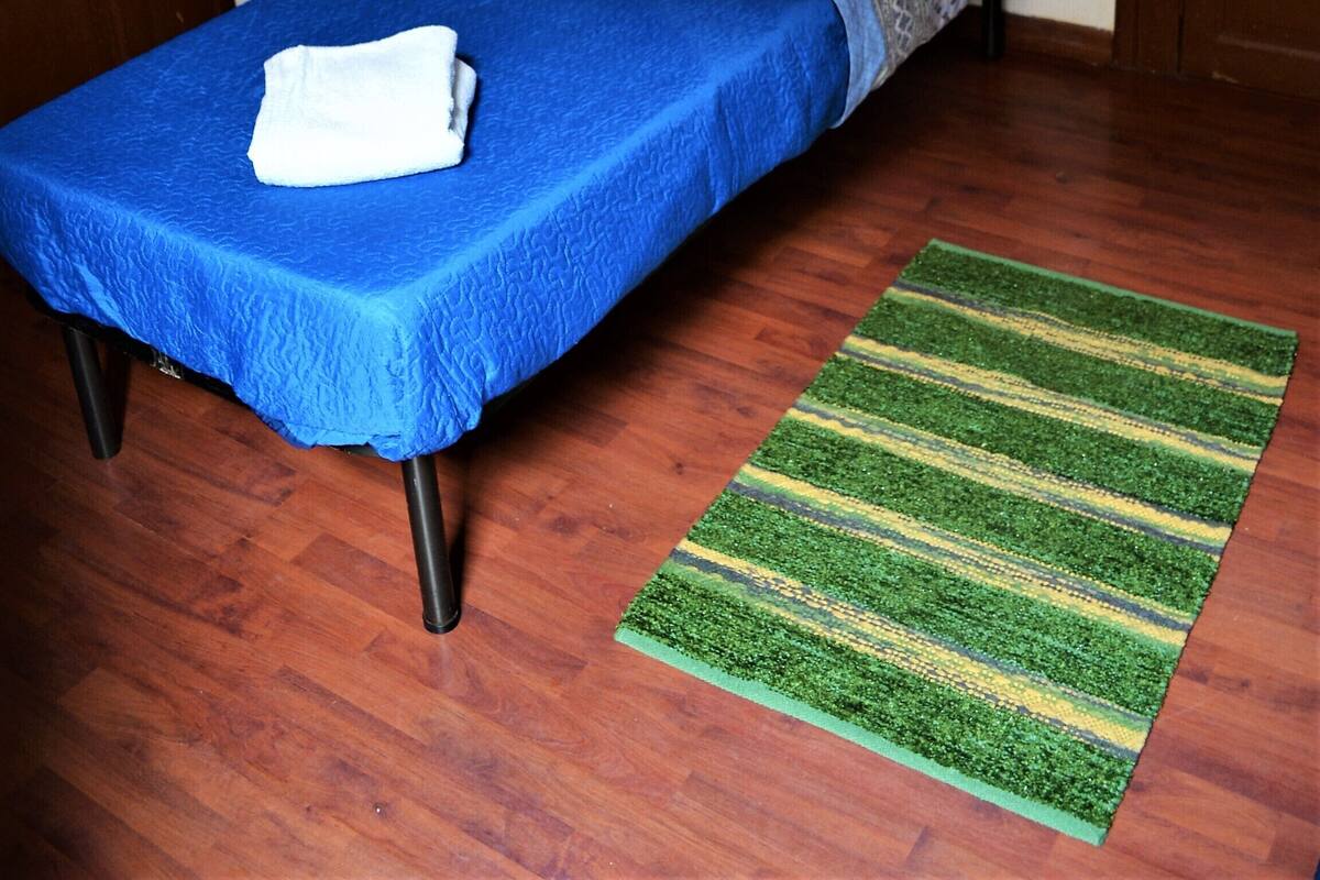 Cama de casal com edredom azul e toalha branca, almofada estampada, pequena prateleira de madeira com objeto decorativo e tapete listrado verde sobre o piso de madeira.