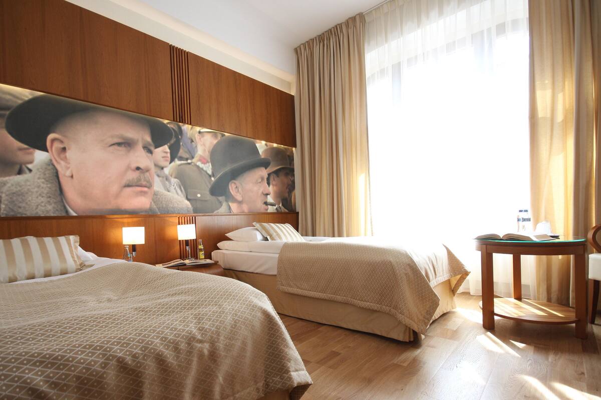 Quarto de hotel com duas camas, uma televisão de parede grande, uma mesinha com um livro e uma garrafa, e uma janela com cortinas.