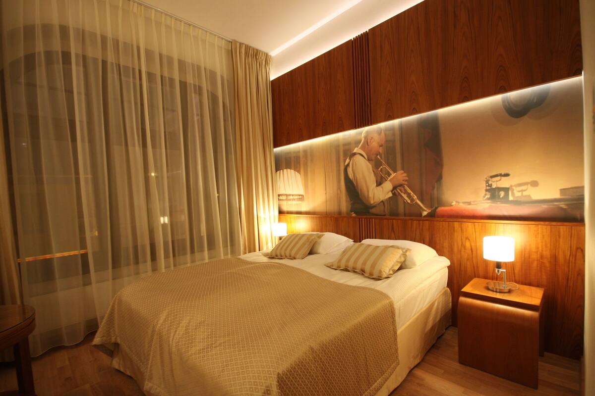 Quarto de hotel com uma cama grande, duas luminárias de cabeceira, um painel de cama de madeira e uma janela ampla com cortinas leves.