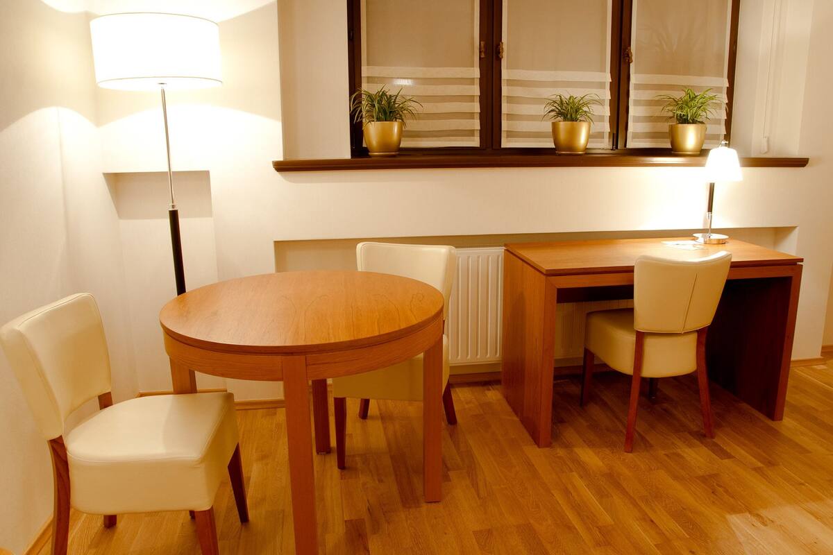 Uma área de refeições com uma mesa redonda de madeira, duas cadeiras brancas, uma escrivaninha de madeira e um radiador. Há duas plantas em vasos no parapeito da janela.