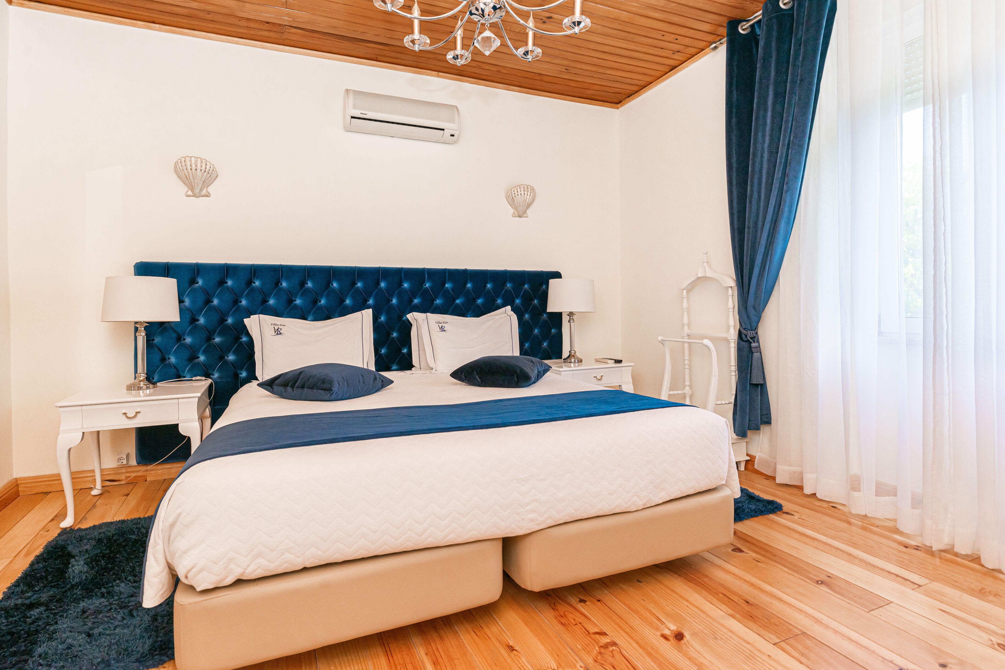 Quarto com cabeceira estofada, cama com roupa de cama branca e azul, duas mesas de cabeceira com abajures, piso de madeira e um ventilador de teto.