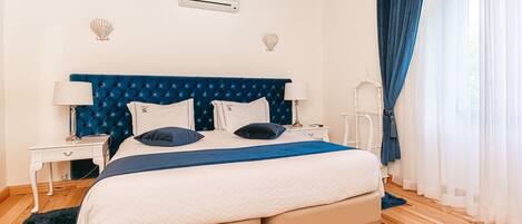 Quarto com cabeceira estofada, cama com roupa de cama branca e azul, duas mesas de cabeceira com abajures, piso de madeira e um ventilador de teto.