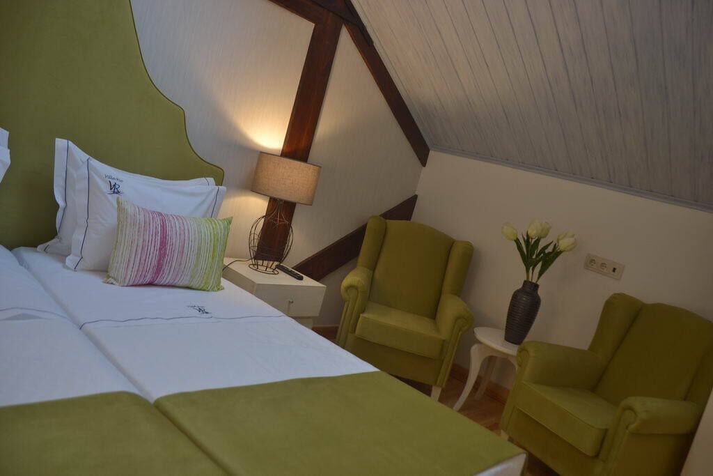 Quarto de hotel com uma cama, duas poltronas verdes, uma mesinha com um vaso de flores e um abajur.