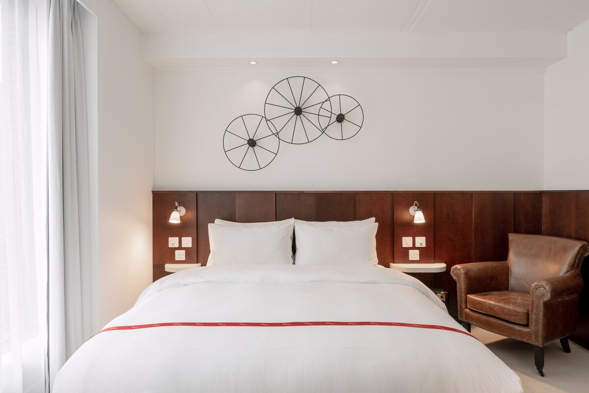Quarto de hotel com uma cama grande, cabeceira de madeira, duas mesas de cabeceira com luminárias, uma poltrona de couro marrom e um aplique circular fixado na parede.