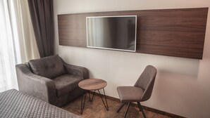 Quarto de hotel moderno com uma cama grande, um sofá cinzento, uma poltrona menor, uma mesa de madeira redonda e uma televisão de tela plana fixada em um painel de madeira.