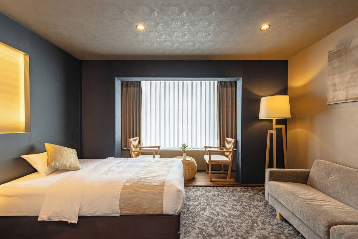 Quarto de hotel moderno com uma cama grande, duas cadeiras, um sofá, uma luminária e uma janela com persianas.