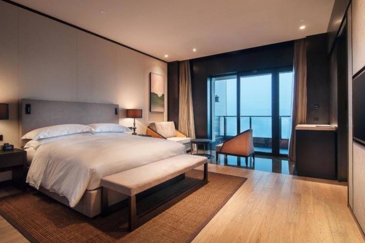 Quarto de hotel moderno com uma cama grande, um banco, uma escrivaninha e vista para a cidade.