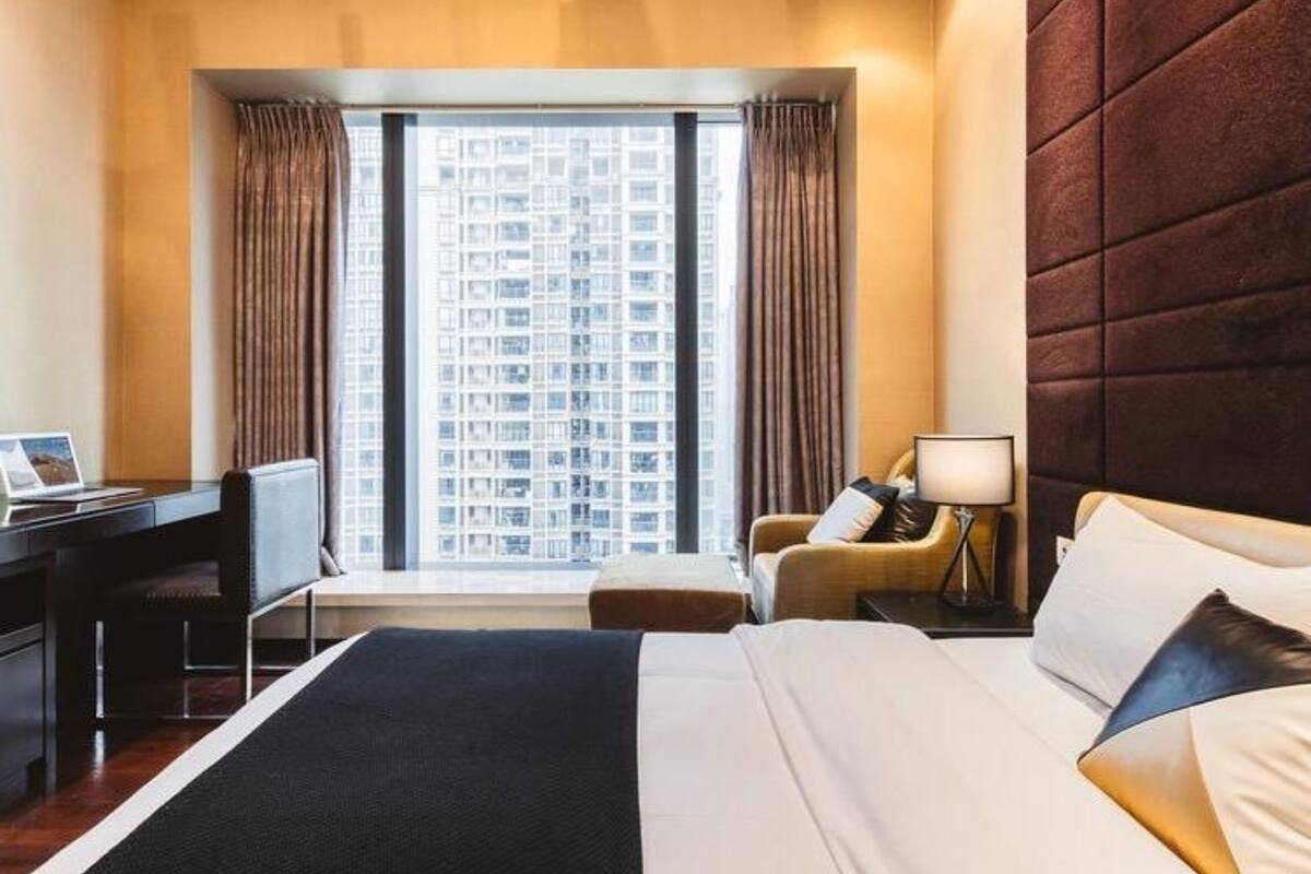 Quarto de hotel com uma cama grande, uma escrivaninha, uma cadeira e vista para um arranha-céu pela janela.