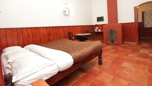 Uma cama de solteiro com roupa de cama branca e castanha, cabeceira de madeira, uma mesa pequena com cadeiras, um relógio de parede e uma lareira.