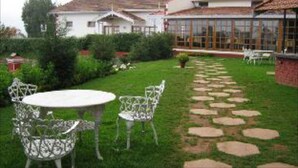 Um jardim com um caminho pavimentado, uma mesa branca com cadeiras e um edifício com telhado em azulejos.