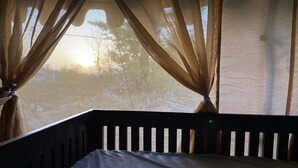 Quarto com cama, cortinas leves e vista para árvores pela janela.