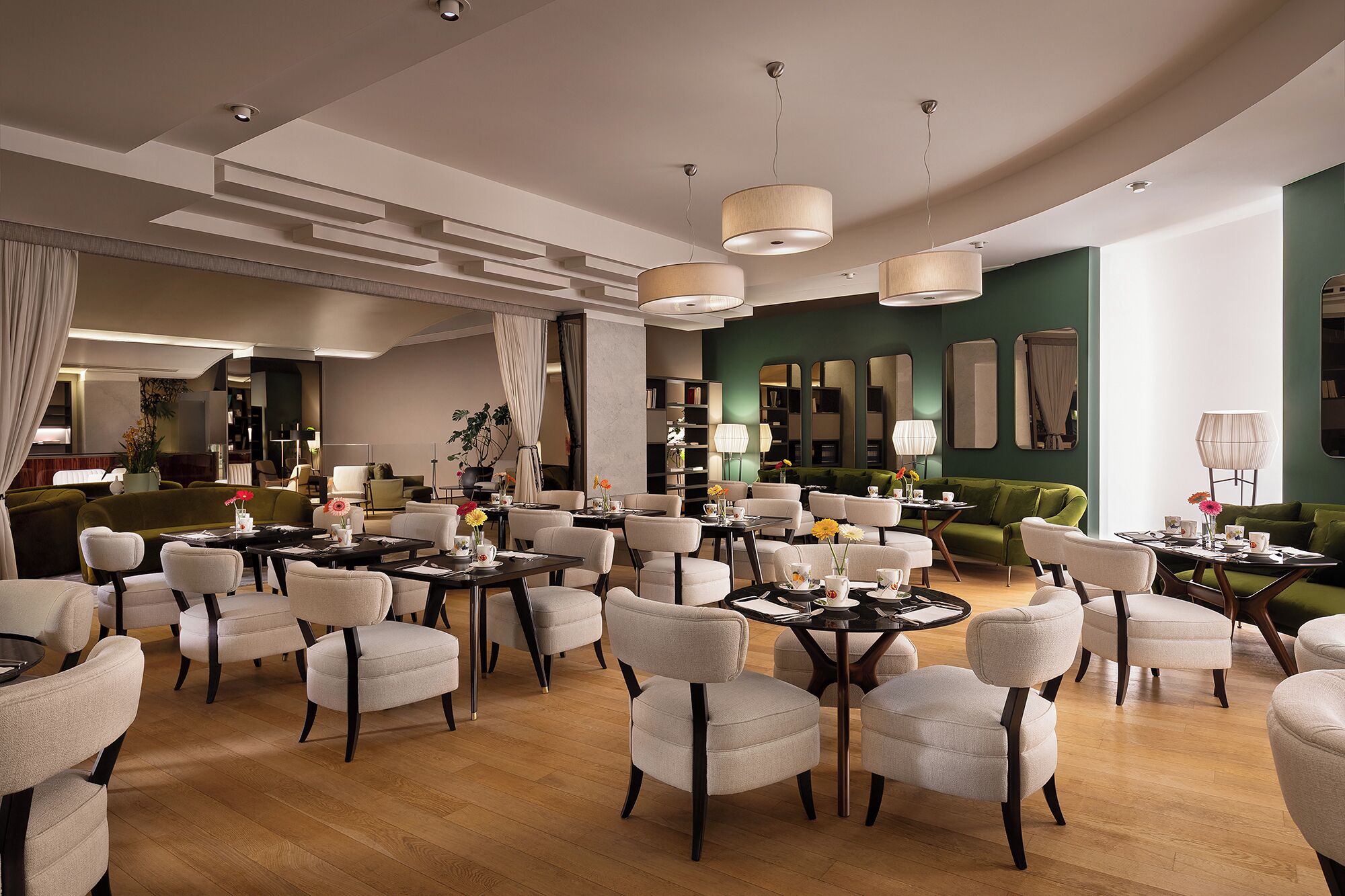 Um amplo hall de hotel com várias áreas de estar, incluindo mesas redondas e sofás, e uma parede verde como detalhe.