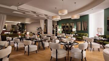 Um amplo hall de hotel com várias áreas de estar, incluindo mesas redondas e sofás, e uma parede verde como detalhe.