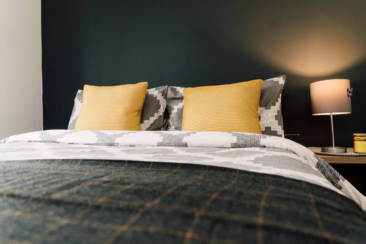 Cama bem arrumada com roupa de cama estampada, parede escura e abajur.