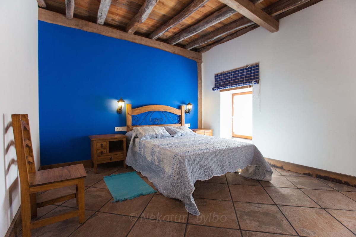 Quarto com cama de madeira, mesa de cabeceira, cadeira e janela com cortina azul.