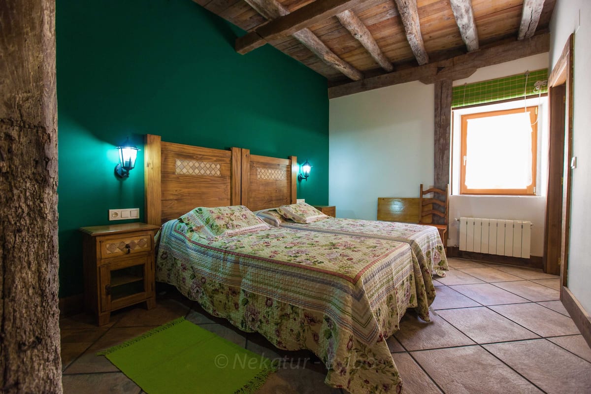 Quarto com duas camas de madeira, uma mesa de cabeceira, um tapete verde e janela com vista.
