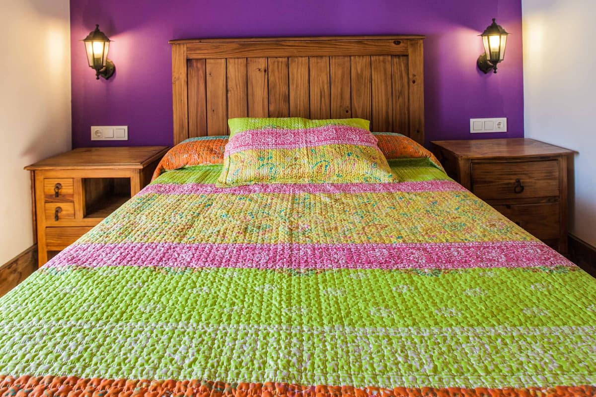 Uma cama com colcha colorida, cabeceira de madeira e duas mesas de cabeceira com luminárias.