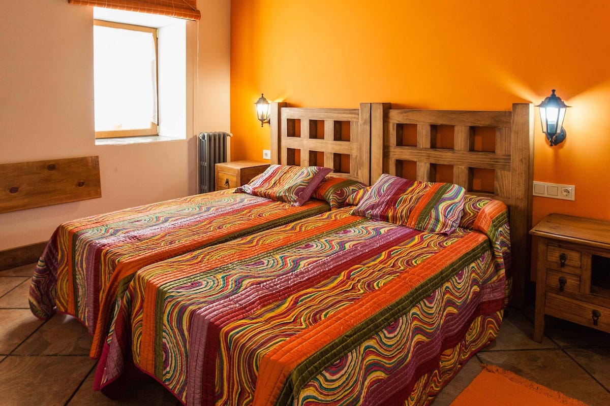 Quarto com teto de madeira, duas camas com roupa de cama colorida, uma janela com persianas e uma mesa de cabeceira com abajur.