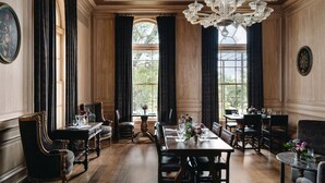 Uma sala de jantar formal com uma longa mesa posta para refeição, ladeada por janelas altas com cortinas, e um lustre acima.