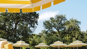 Uma área à beira da piscina com guarda-sóis listrados em amarelo e branco, espreguiçadeiras e uma mesa com uma bebida.