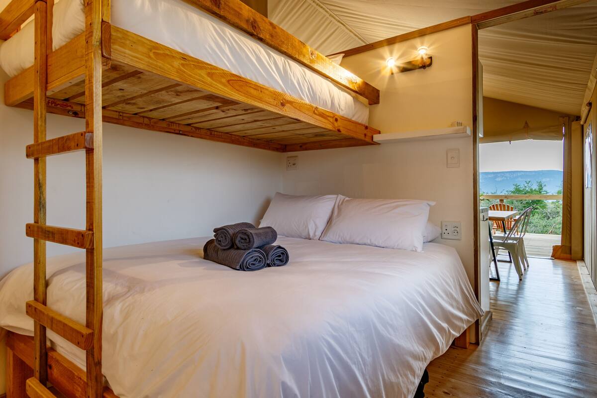 Quarto com beliche, escada de madeira, roupa de cama branca e vista para o exterior.