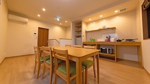 Uma sala de jantar com mesas e cadeiras de madeira, uma área de cozinha com prateleiras e eletrodomésticos, e um piso de madeira.