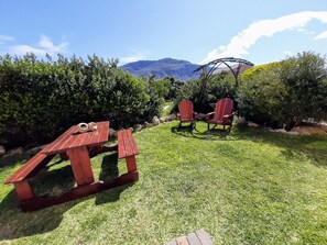 Uma área para piquenique com uma mesa de madeira e duas cadeiras Adirondack vermelhas, em um gramado bem cuidado e cercada por arbustos densos.