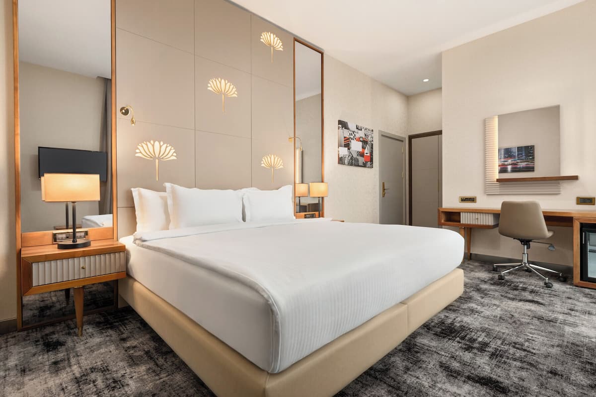 Quarto de hotel com uma cama grande, abajures, uma escrivaninha com cadeira, um espelho e uma obra de arte pendurada na parede.