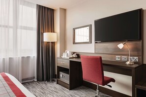 Quarto de hotel com cama, escrivaninha com cadeira, televisão, luminária e janela com cortinas.