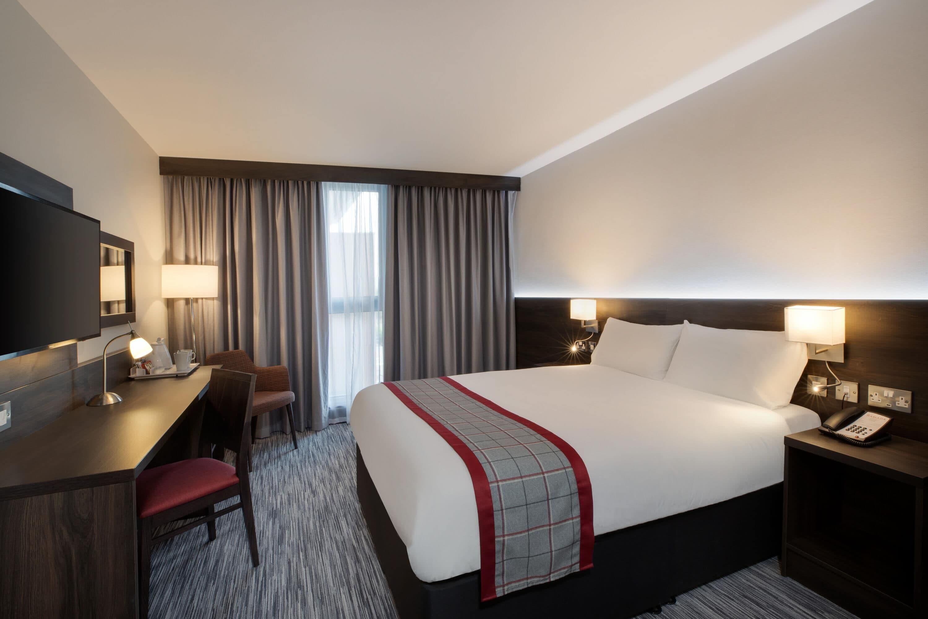 Quarto de hotel com uma cama grande, uma escrivaninha com cadeira, televisão e mesa de cabeceira com telefone.