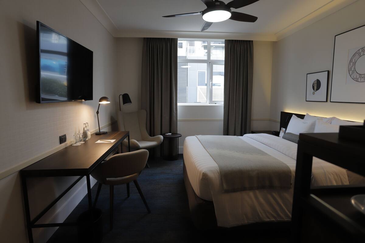 Quarto de hotel com cama, escrivaninha, cadeira, televisão e ventoinha no teto.