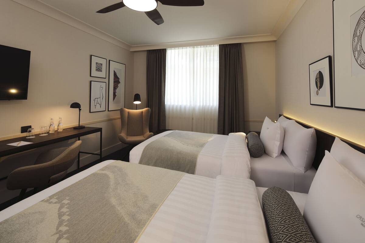 Quarto de hotel com duas camas, uma escrivaninha, uma cadeira, televisão e obras de arte nas paredes.