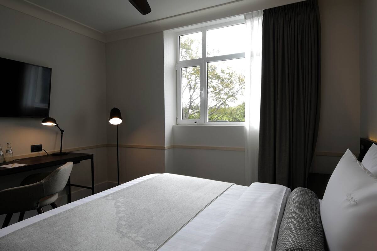 Quarto de hotel com cama, escrivaninha, cadeira, televisão e janela com cortinas.