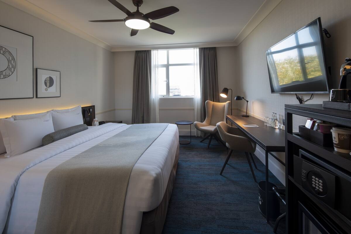 Quarto de hotel com uma cama grande, uma escrivaninha com cadeira, uma televisão e um ventilador de teto.