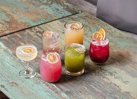 Cinco copos de bebidas de cores variadas sobre uma mesa de madeira rústica.