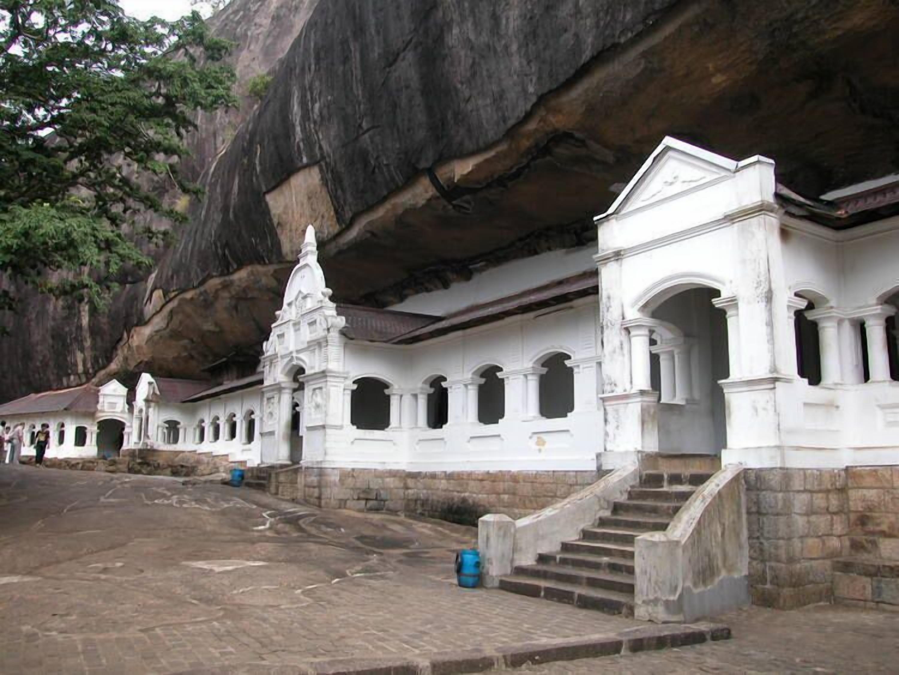 Um edifício branco com uma escadaria que leva a uma estrutura semelhante a um templo, diante de uma grande formação rochosa.