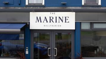 Um edifício com uma placa que diz "The Marine Ballybunion" e outra placa com a inscrição "OH! I DO LIKE TO BE BESIDE THE SEASIDE".