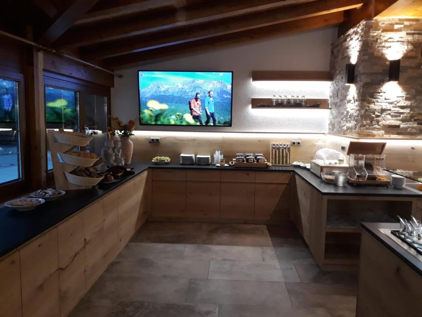 Uma cozinha moderna com parede decorada em pedra, TV de tela plana e diversos itens de café da manhã sobre o balcão.