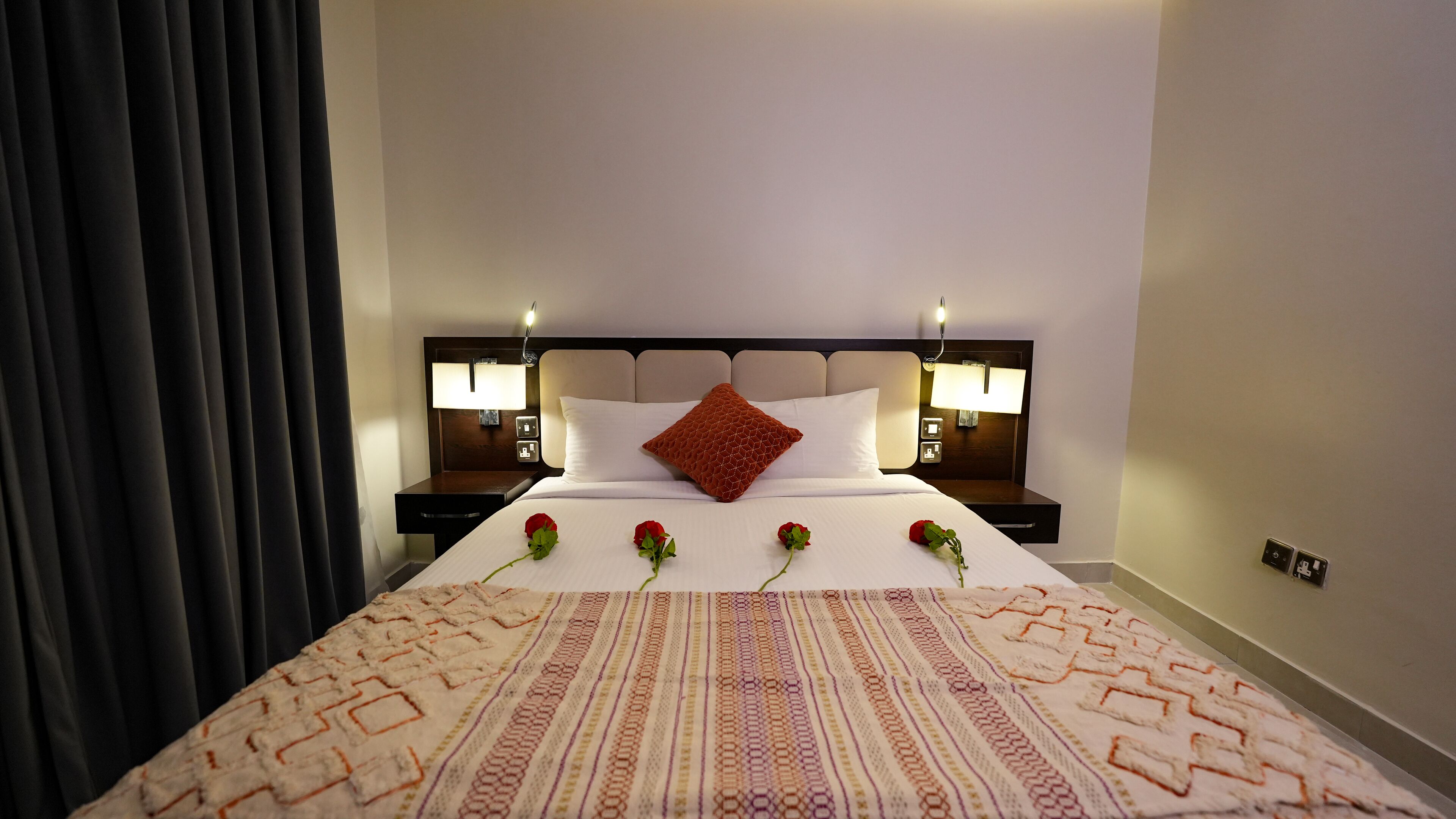 Quarto de hotel com cama bem arrumada, duas luminárias de cabeceira, cabeceira com iluminação integrada e uma janela grande com cortinas.