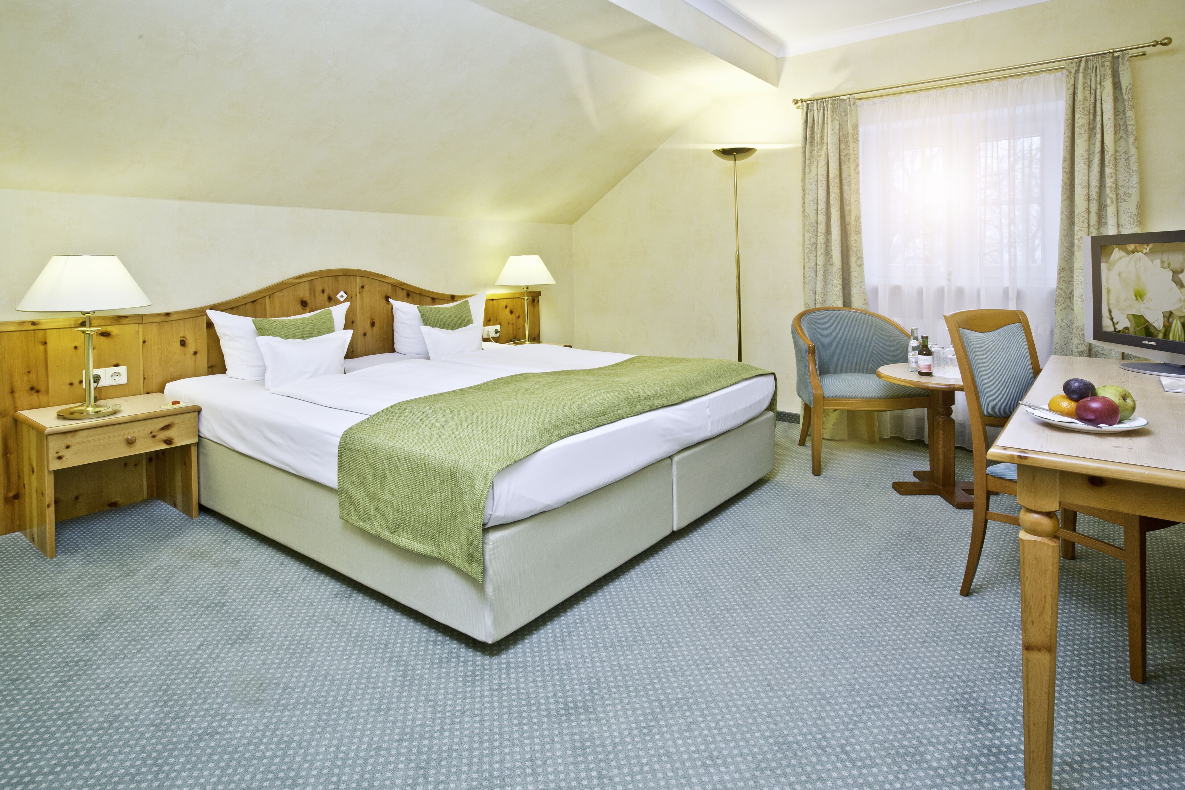 Quarto de hotel com cama, mesas de cabeceira, escrivaninha, cadeira e televisão.