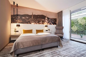 Quarto de hotel moderno com uma cama grande, um mural com vista para a cidade na parede, vegetação visível pela janela e uma poltrona confortável.